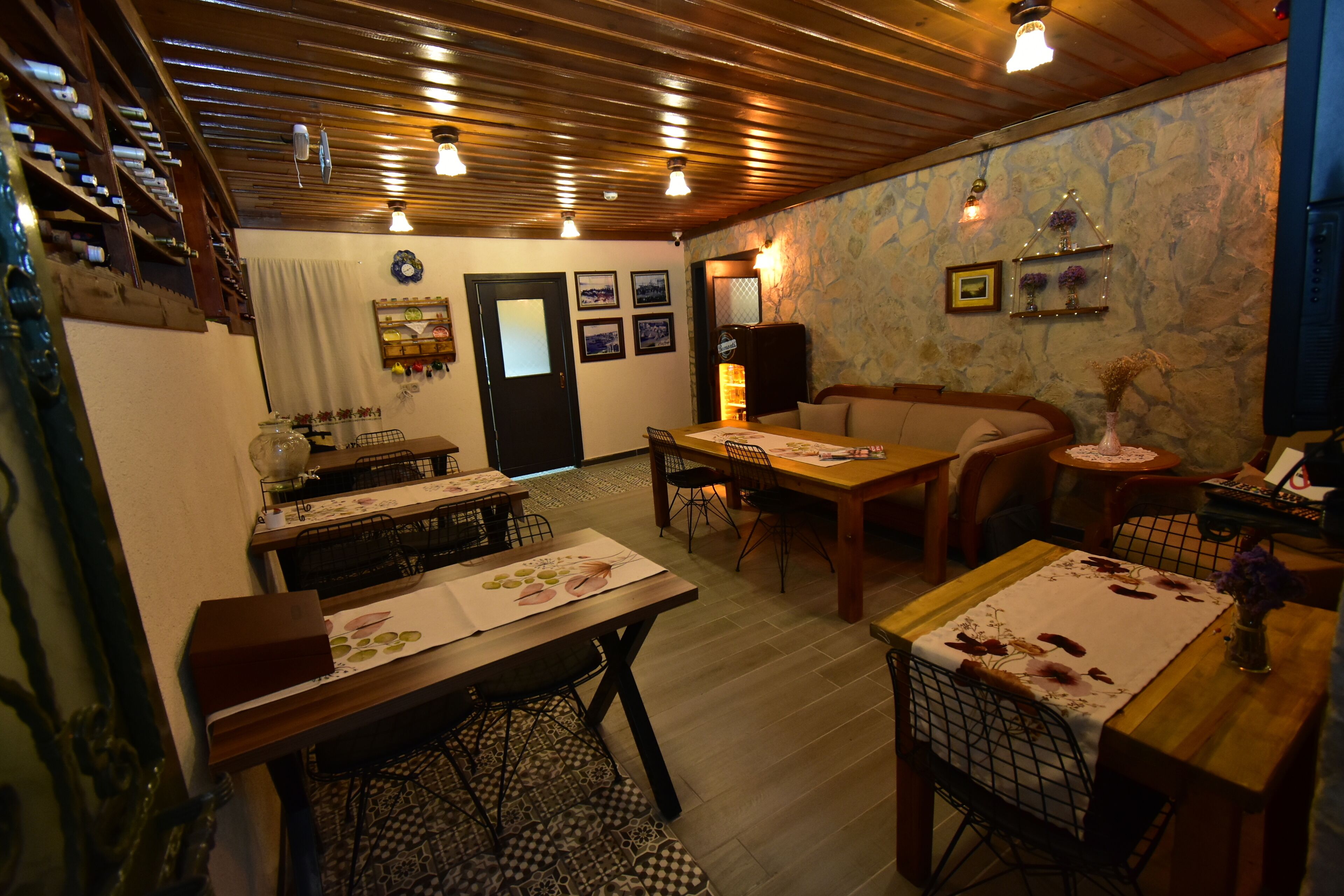 Uma área de refeições acolhedora com mesas e cadeiras de madeira, um espaço de bar com garrafas e uma parede de pedra decorada com quadros.