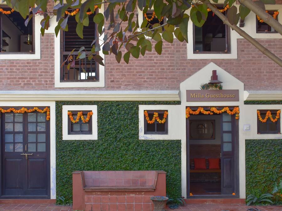 Um edifício de tijolos com uma placa que diz "Milla Guesthouse". Possui uma porta de madeira escura, janelas com detalhes laranja e um banco na frente.