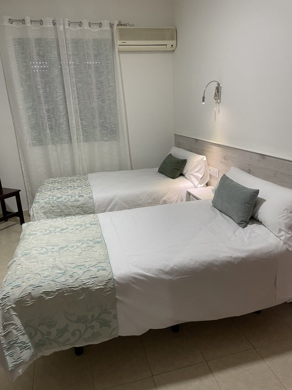 Quarto com duas camas, mesa de cabeceira, ar condicionado na parede e janela com cortina.