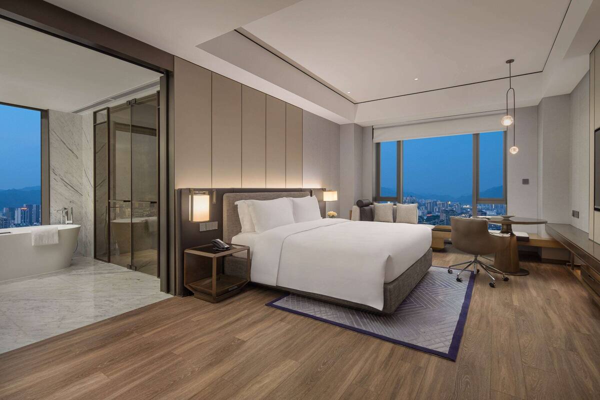 Quarto de hotel moderno com uma cama grande, uma escrivaninha, uma cadeira, banheiro com banheira e vista para a cidade através de amplas janelas.