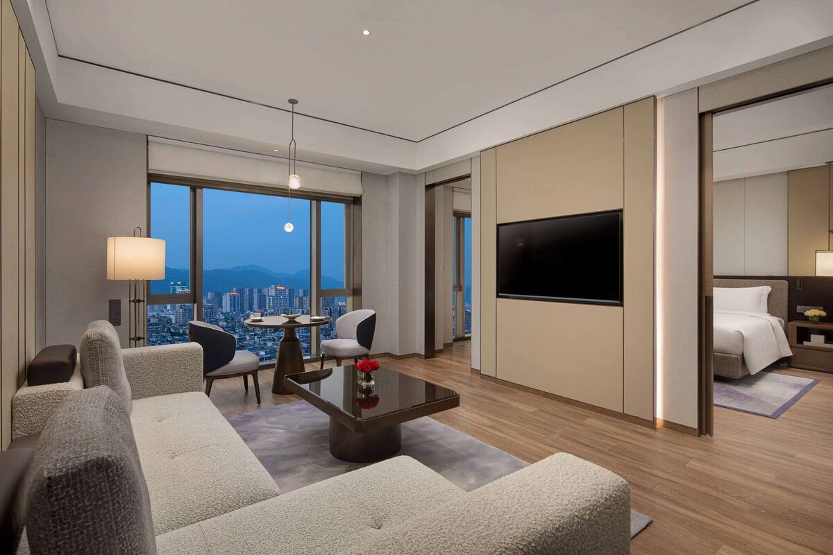 Quarto de hotel moderno com vista para a cidade, uma TV de tela plana, um sofá, uma mesa de centro e um espaço de refeições com uma luminária.