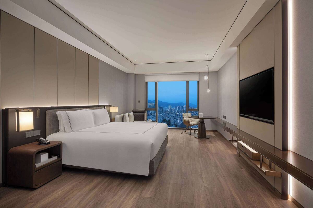 Quarto de hotel moderno com uma cama grande, uma escrivaninha, uma TV e vista para a cidade.