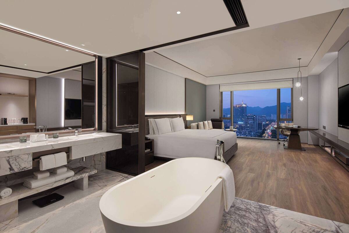 Quarto de hotel moderno com uma cama grande, uma banheira, toucador com espelho e vista para a cidade.