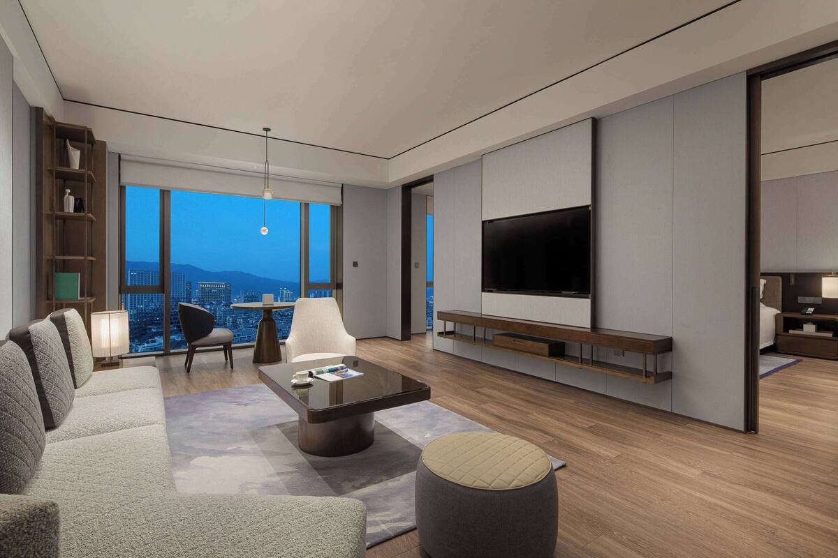 Sala de estar moderna com sofá, mesa de centro e televisão. Vista urbana visível por grandes janelas.