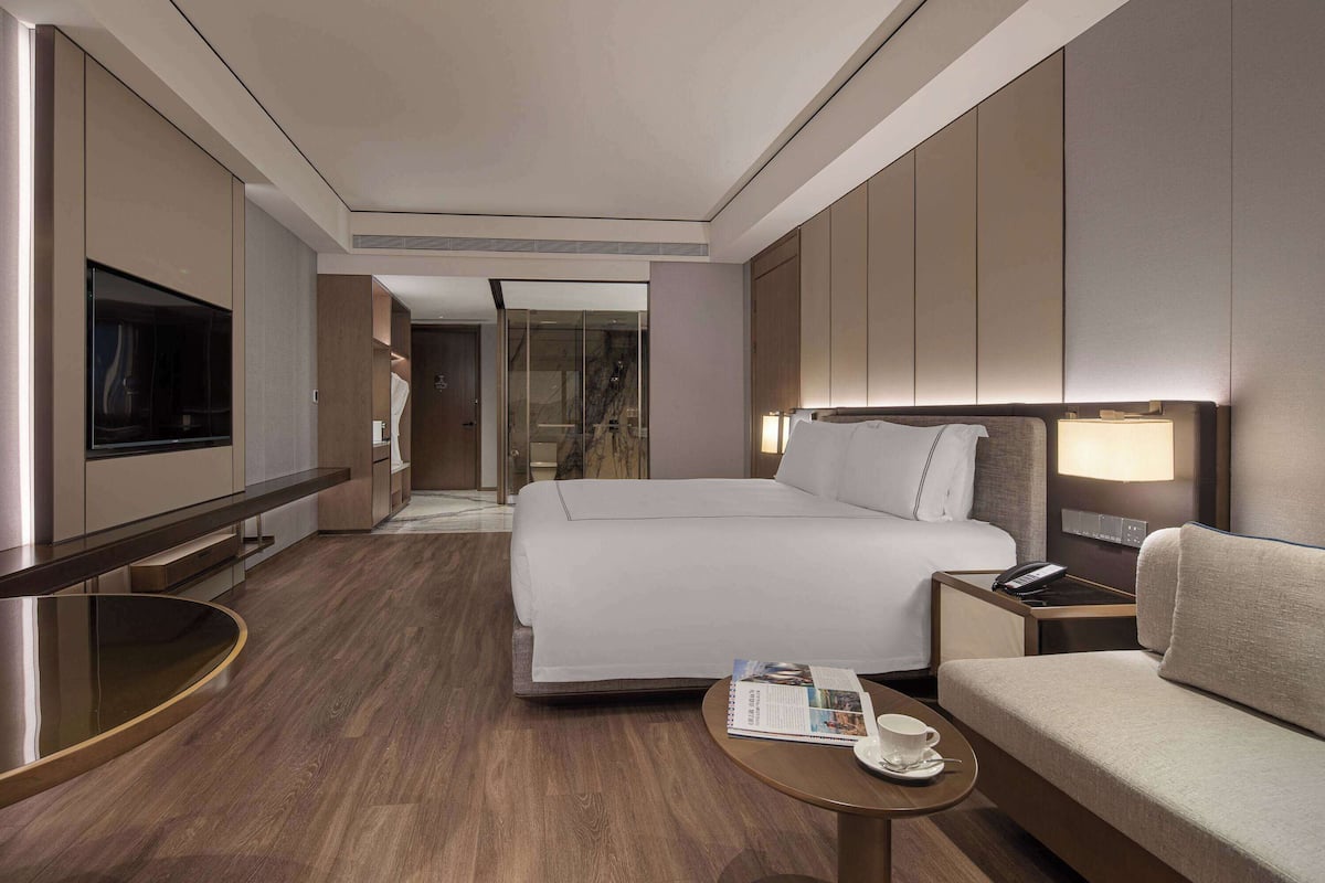 Quarto de hotel moderno com uma cama grande, um sofá, uma mesinha, uma televisão e uma porta de vidro que dá para um banheiro.