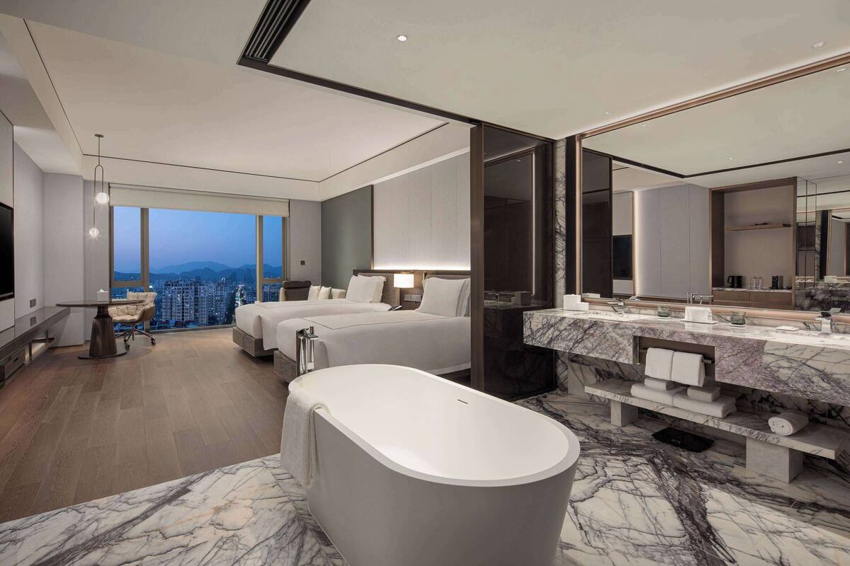 Quarto de hotel moderno com banheira grande, cama de casal, sofá, escrivaninha e vista para a cidade.