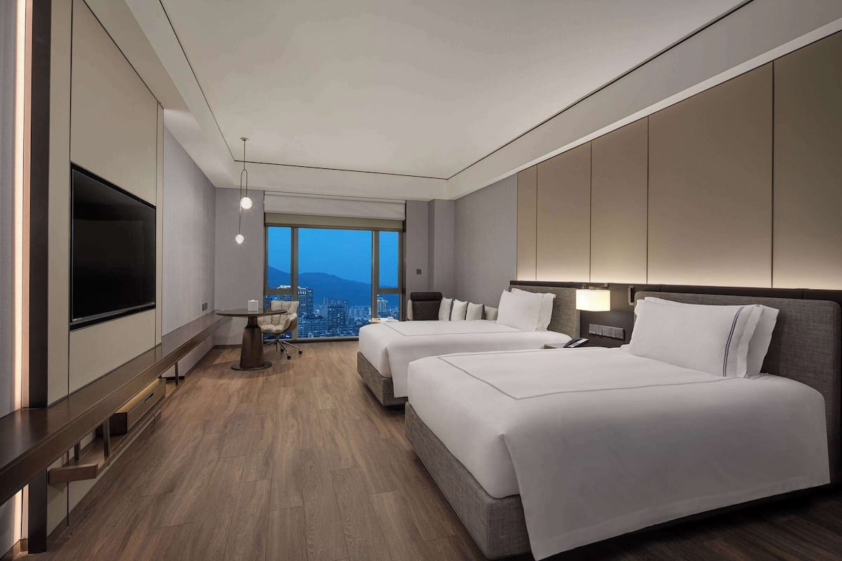 Quarto de hotel com duas camas, televisão, escrivaninha e vista para a cidade.
