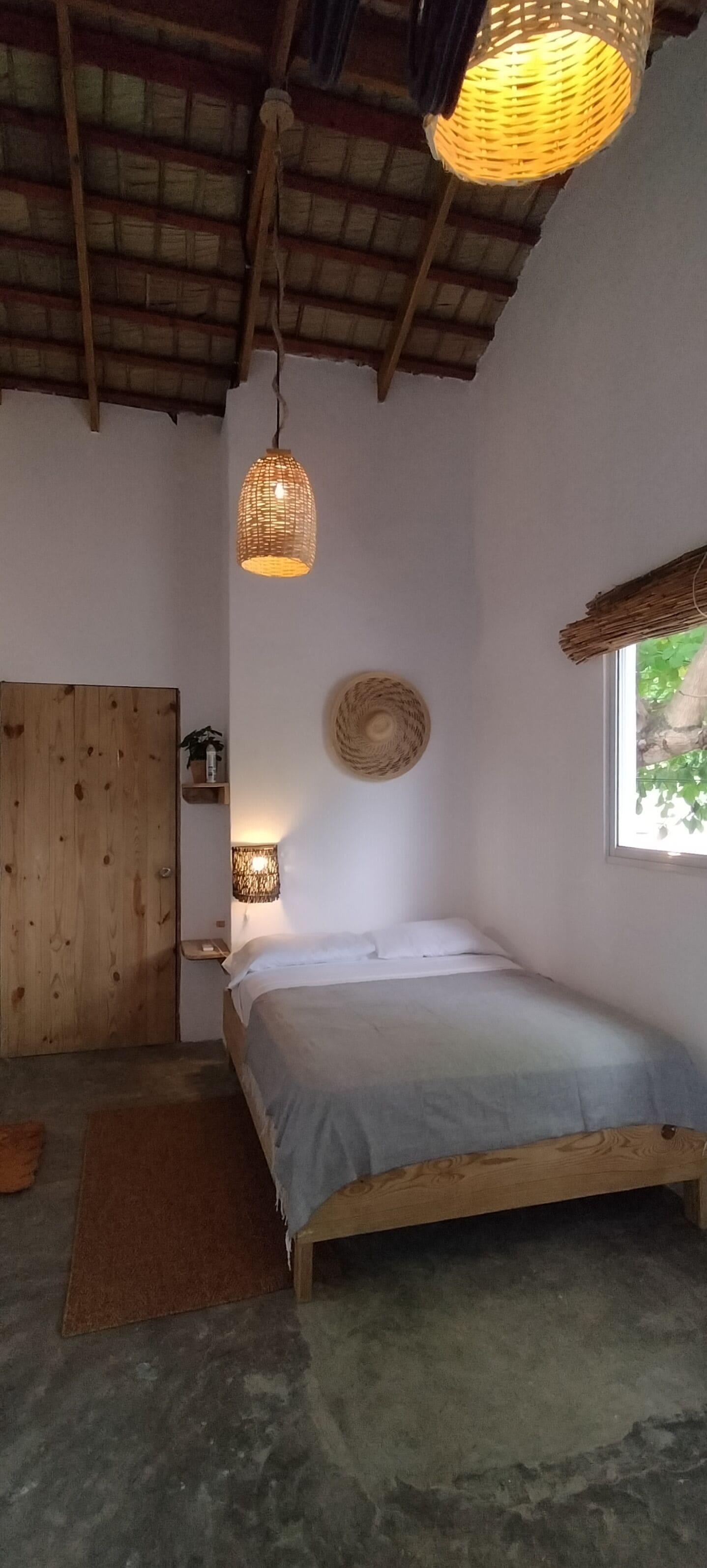 Quarto com cama de madeira, um pendão de parede tecido, luminária tecida e janela com vista para vegetação.