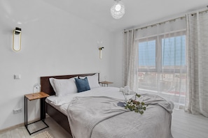 Quarto moderno com cama de madeira, roupa de cama branca, mesa de cabeceira com abajur e vista da cidade através de cortinas leves.