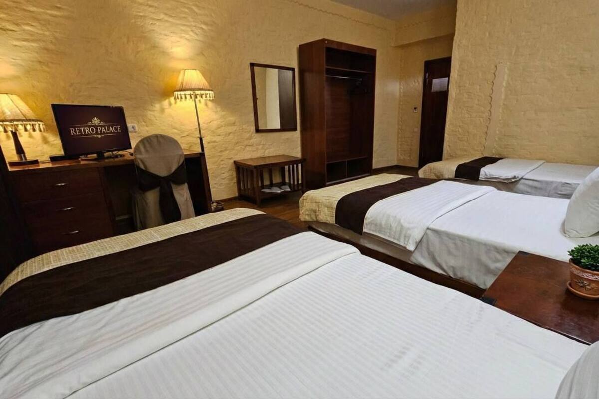Quarto de hotel com duas camas, uma escrivaninha, uma cadeira, um guarda-roupa e uma pequena mesa com uma planta.