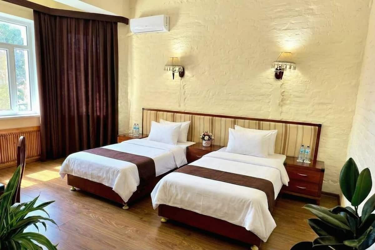 Quarto de hotel com duas camas, piso de madeira, janela com cortinas, luminárias fixadas na parede e um radiador.