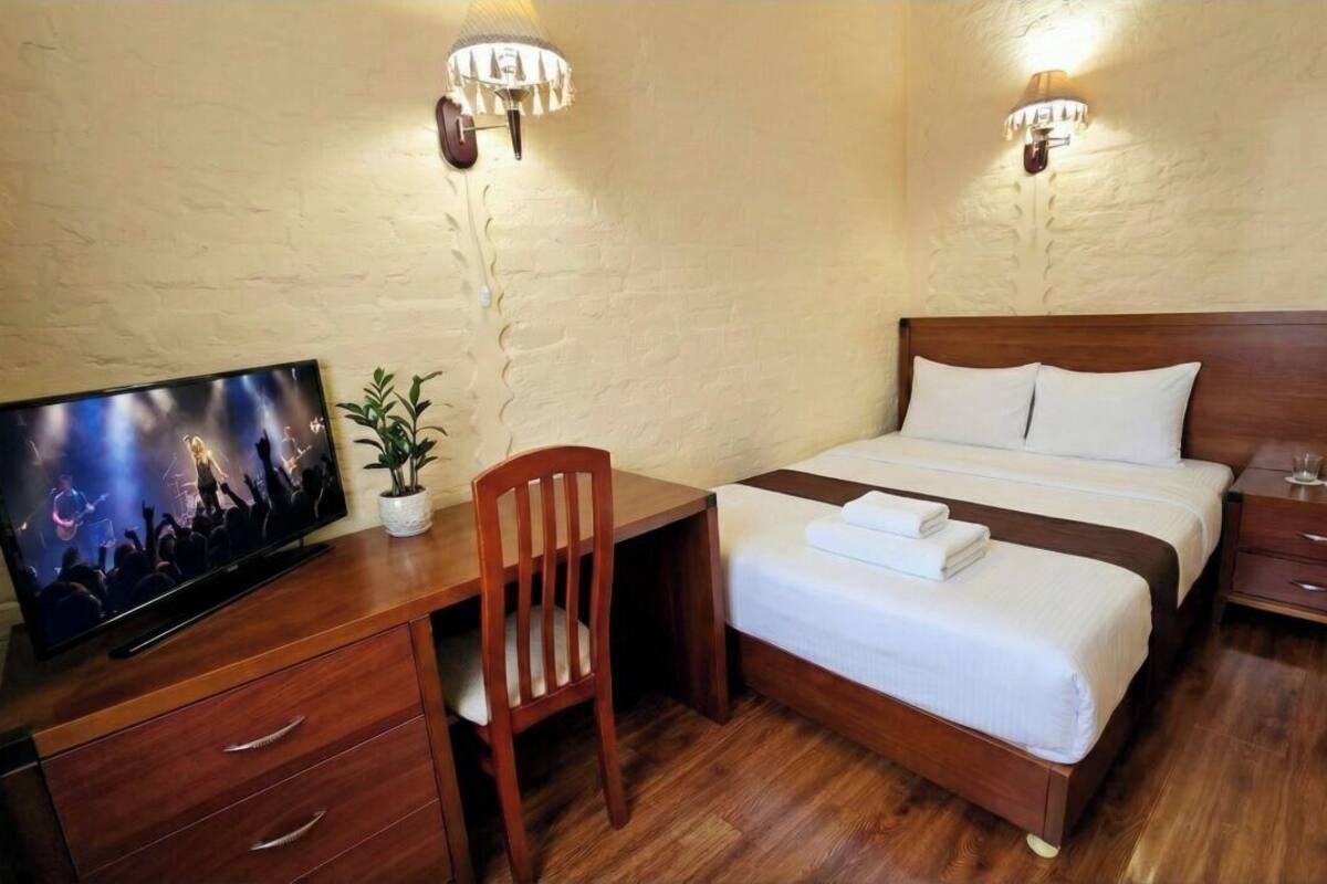 Quarto de hotel com uma cama, mesas de cabeceira, escrivaninha com uma cadeira, televisão e duas luminárias fixadas na parede.