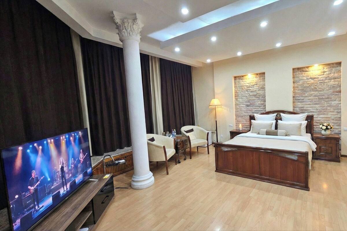 Quarto de hotel com uma cama grande, televisão, escrivaninha e duas luminárias.