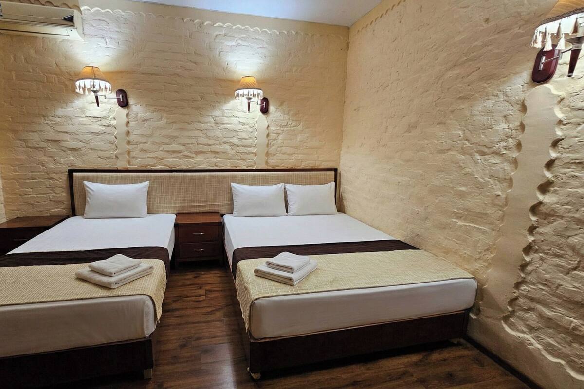 Quarto com duas camas, cada uma com travesseiro e toalha dobrada. Há luminárias na parede e um aparelho de ar condicionado.