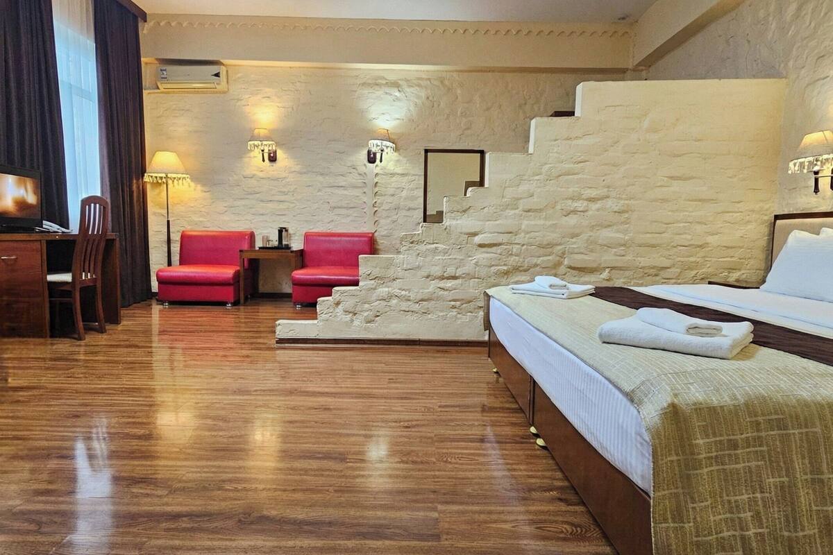 Quarto de hotel com piso de madeira, cama com roupa de cama branca, televisão, escrivaninha e duas cadeiras vermelhas.