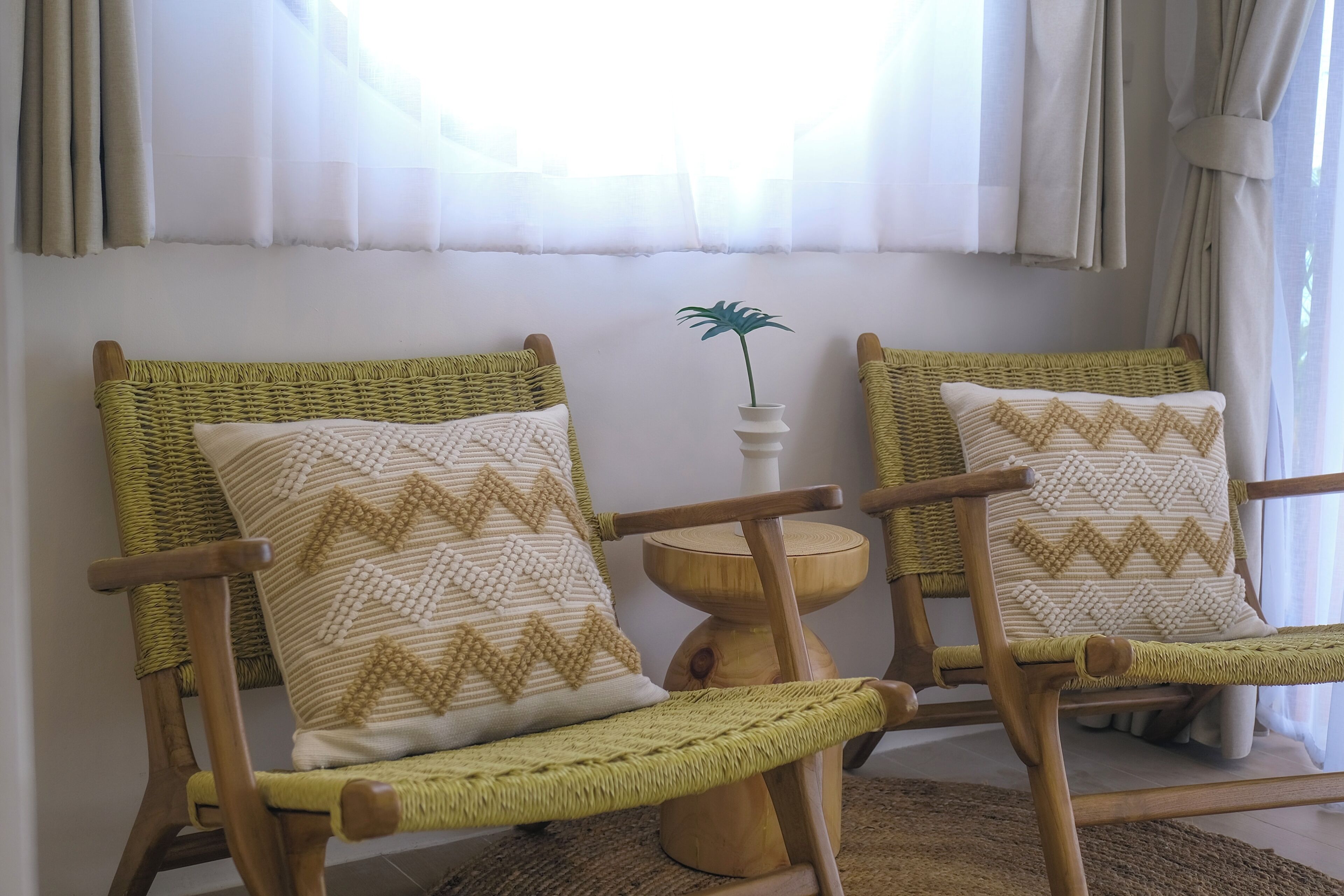 Duas cadeiras de vime com almofadas estampadas, uma pequena mesa de madeira com uma planta e uma janela com cortinas leves.