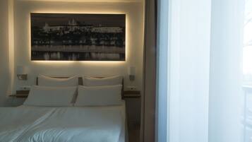 Quarto de hotel com uma cama, duas mesas de cabeceira e um quadro na parede retratando uma paisagem urbana.