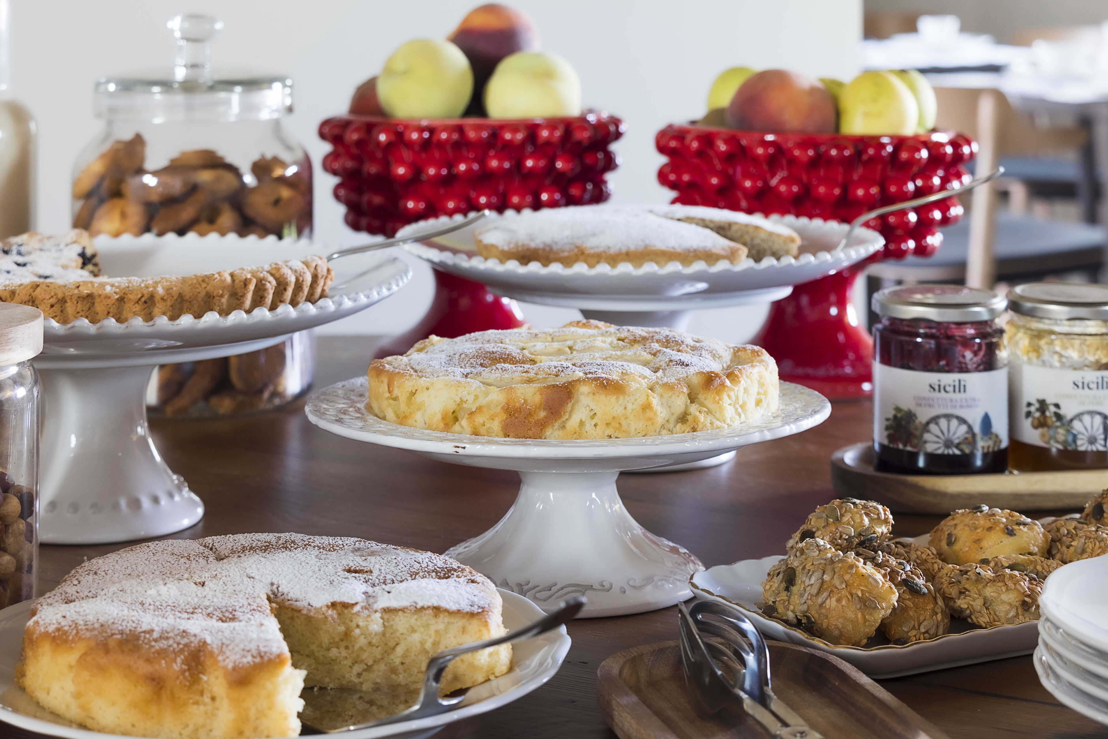 Diversas sobremesas em travessas brancas e vermelhas, incluindo bolos e biscoitos, com potes de geleia e uma tigela de frutas ao fundo.
