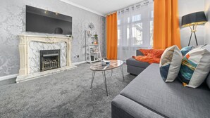 Sala de estar com lareira, sofá cinzento, cortinas laranja e luminária.