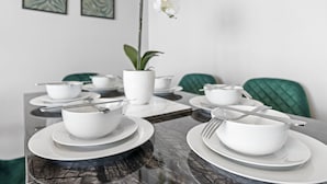 Mesa de jantar posta com tigelas, pratos e talheres brancos, acompanhada por um vaso de planta e cadeiras verdes.