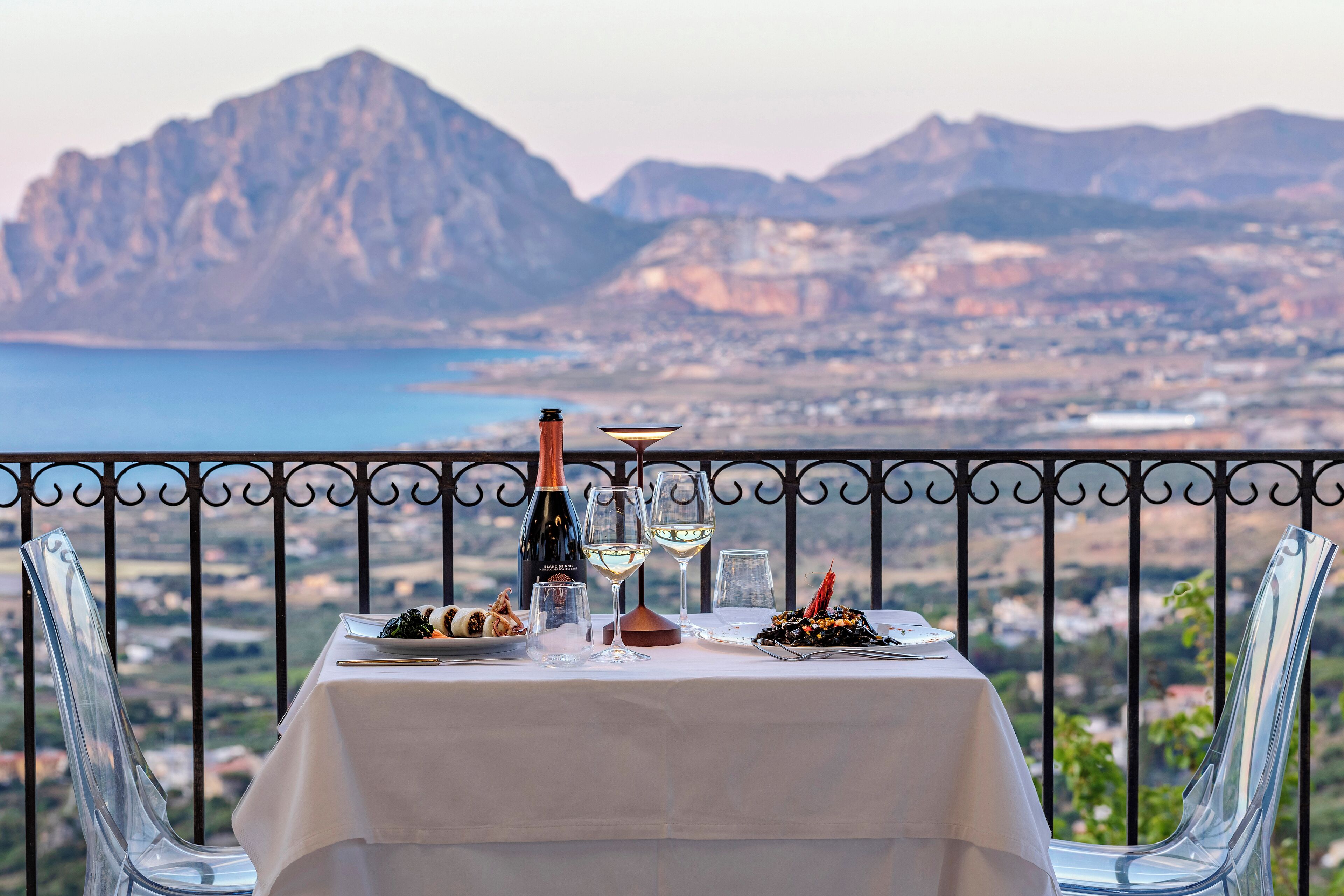 Mesa posta com copos de vinho, uma garrafa e um prato de comida numa varanda com vista para as montanhas.