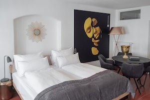 Quarto de hotel moderno com uma cama grande, uma mesa redonda com cadeiras, um abajur e um objeto decorativo na parede.
