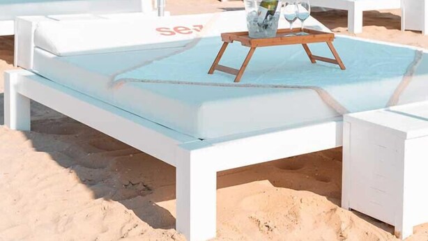 Uma área de descanso à beira-mar, com cadeiras e guarda-sóis brancos, piscina e um bar com mesas ao ar livre.