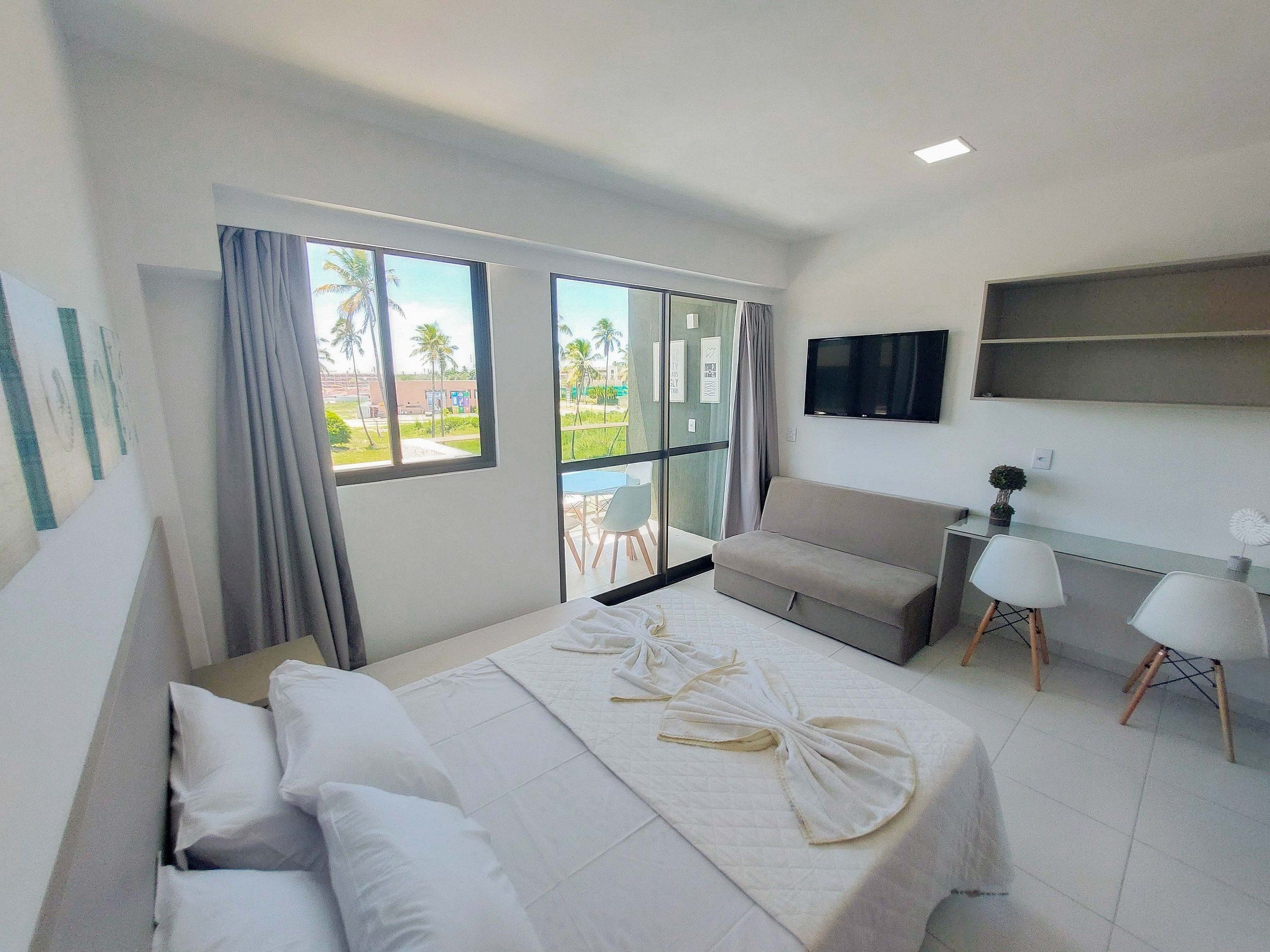 Quarto de hotel com cama, sofá, escrivaninha, televisão e janelas amplas com vista para palmeiras.