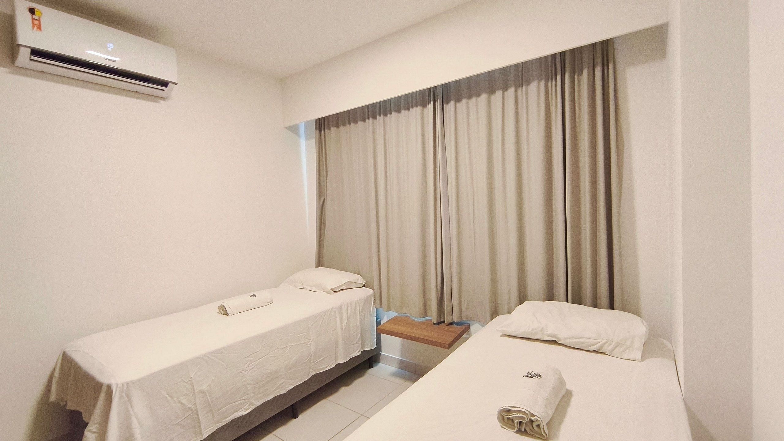 Quarto de hotel com duas camas, uma janela com cortinas, um ar condicionado e uma mesa de cabeceira de madeira.
