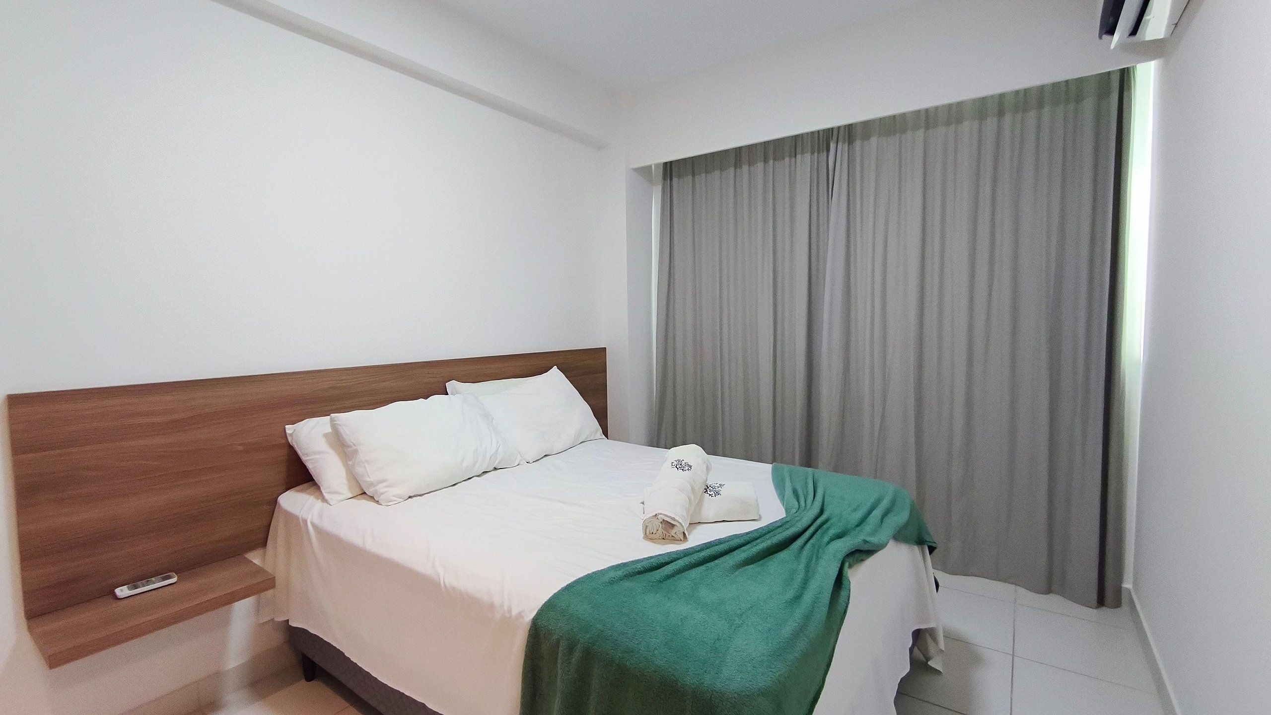 Quarto de hotel com cabeceira de madeira, cama com roupa de cama branca, manta verde e janela com cortinas cinzentas.