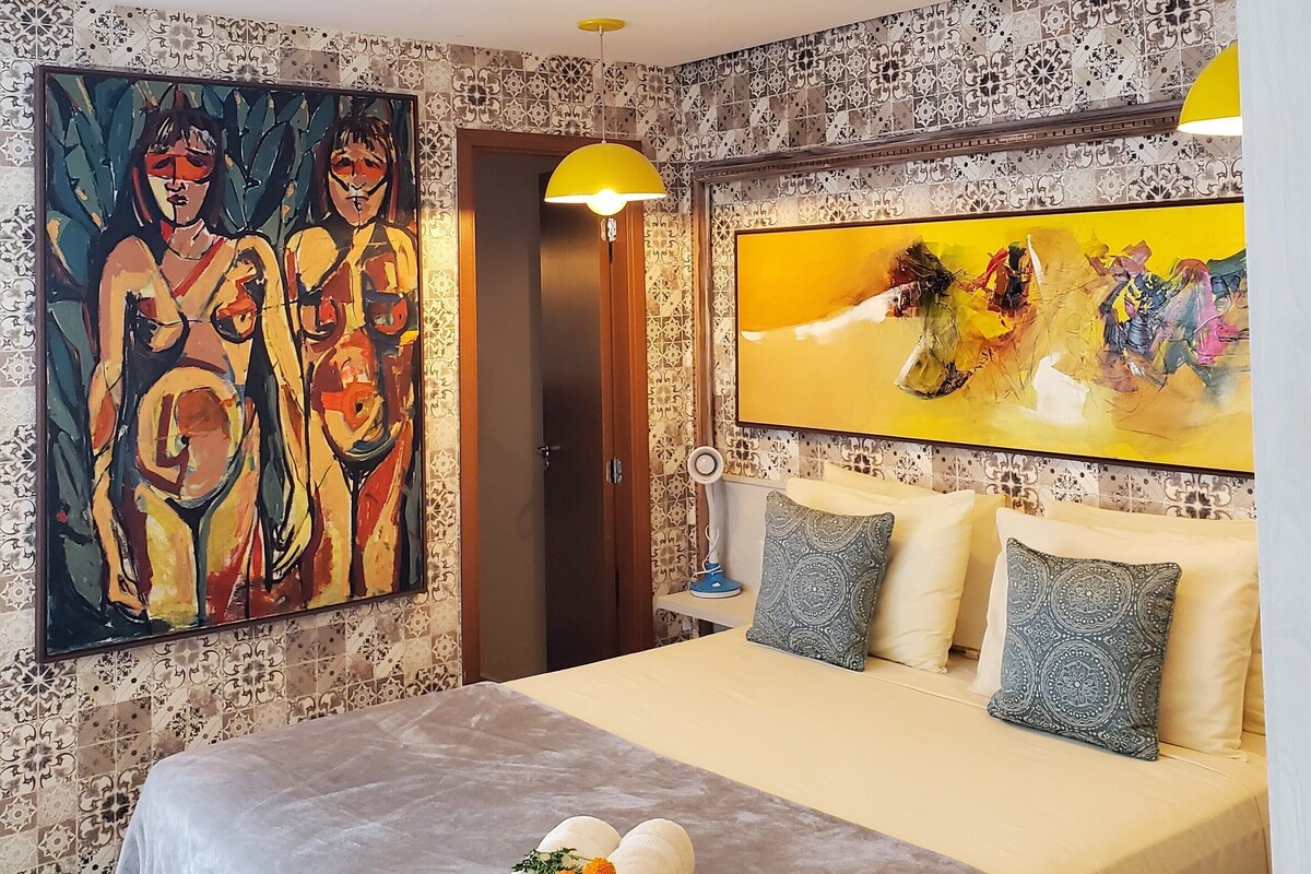 Quarto de hotel com cama, obras de arte nas paredes, ventoinha no teto e uma janela grande.