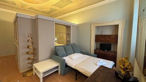 Quarto compacto com cama, mesinha, sofá, TV e teto com detalhes decorativos.