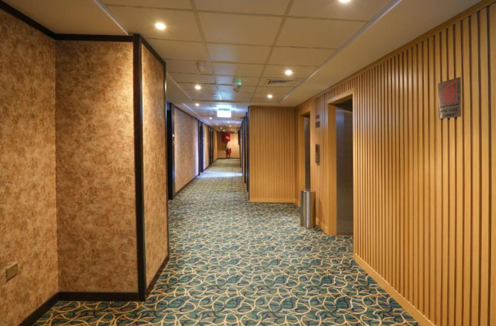 Corredor de hotel com carpete estampado, painéis de madeira e armário de mangueira de incêndio.