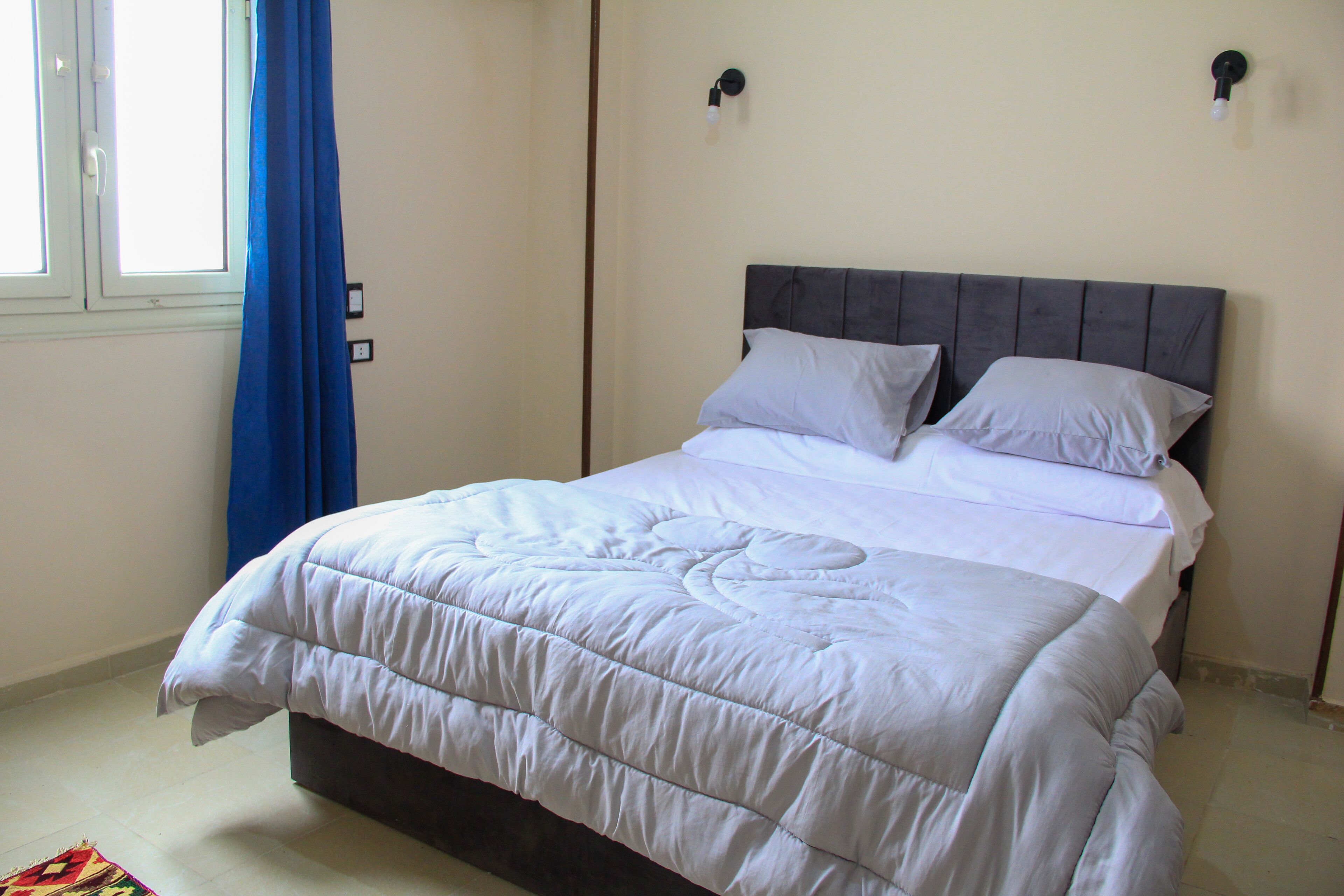 Cama bem arrumada com um edredom branco e almofadas, cabeceira escura e cortina azul parcialmente recolhida.
