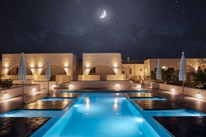 Uma área de piscina à noite, com várias piscinas iluminadas por luzes no teto, e uma lua crescente visível no céu.