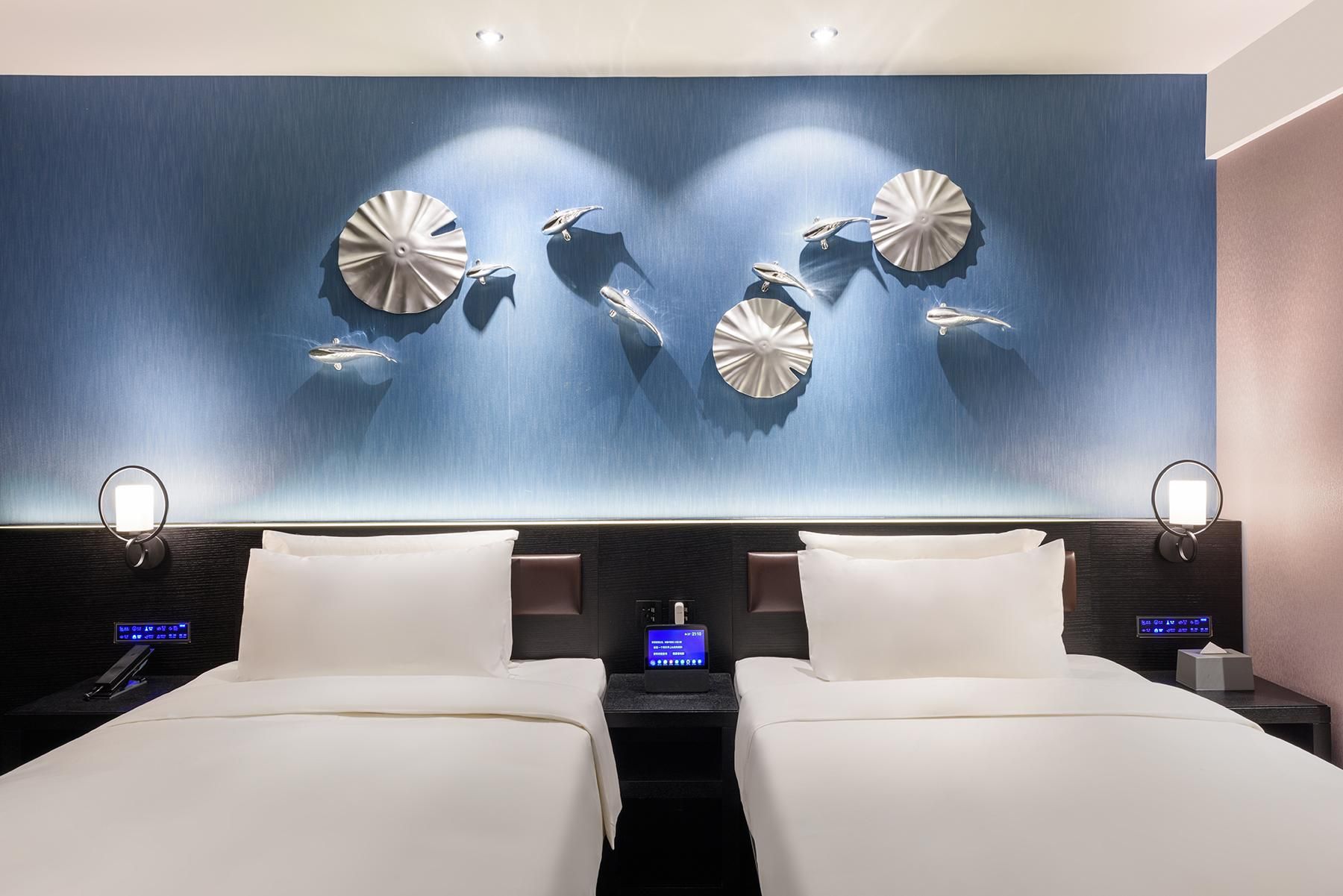 Quarto de hotel com duas camas, luminária fixada na parede e relógio digital na mesa de cabeceira.