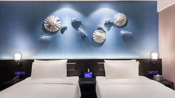 Quarto de hotel com duas camas, luminária fixada na parede e relógio digital na mesa de cabeceira.
