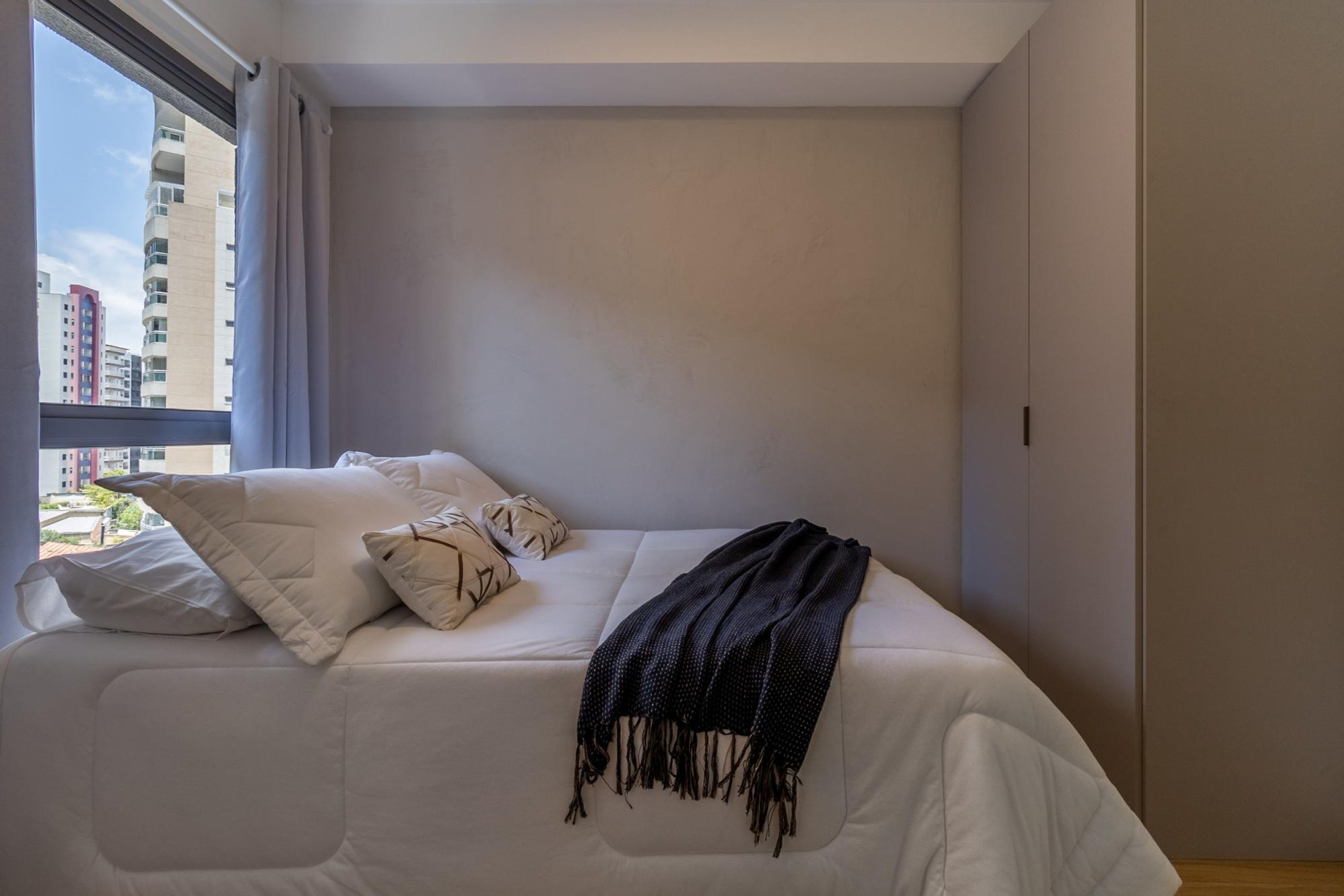 Quarto com cama, travesseiros, cobertor e janela com vista para edifícios.