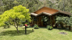 Uma cabana rústica com telhado inclinado, cercada por vegetação exuberante e um cenário montanhoso.