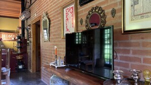 Uma sala de estar acolhedora com parede de tijolos, televisão e diversos objetos decorativos, incluindo quadros e um espelho.