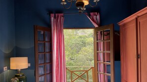 Quarto com uma cama grande, mesa de cabeceira com luminária, guarda-roupa de madeira e vista para um deck de madeira e vegetação através de portas francesas abertas.