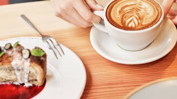 Uma chávena de café com arte de latte num pires, um garfo num prato com uma sobremesa, e uma mesa de madeira.