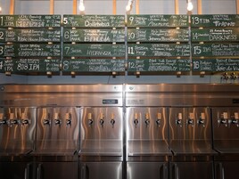 Área de torneiras de cerveja com várias opções e um quadro negro listando os tipos de cerveja e seus preços.