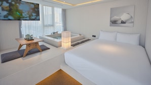 Quarto de hotel moderno com uma cama grande, um pequeno espaço de estar com mesa e cadeiras, uma banheira e uma janela ampla com cortinas.