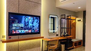 Quarto de hotel moderno com uma grande TV de tela plana na parede, uma escrivaninha de madeira com cadeira, uma mesinha e um espelho.