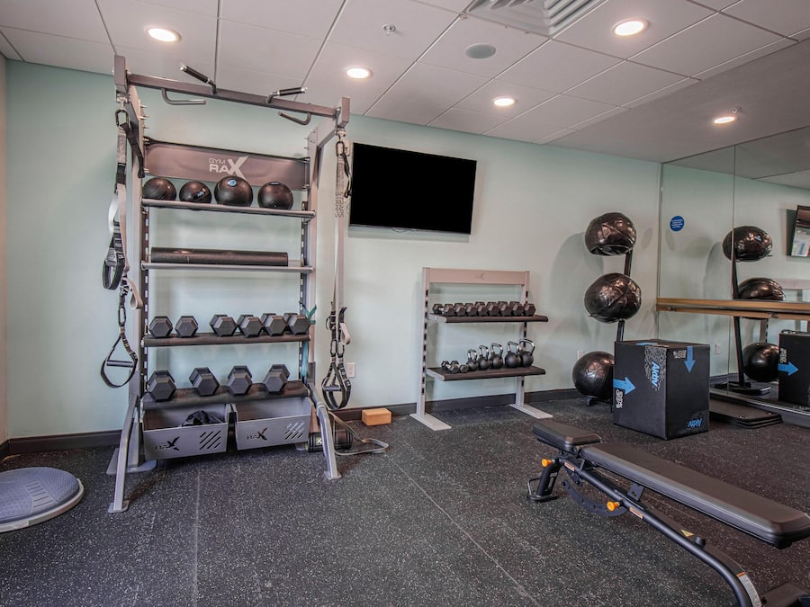 Uma academia com barra de exercícios, halteres, kettlebells, um banco e um espelho.