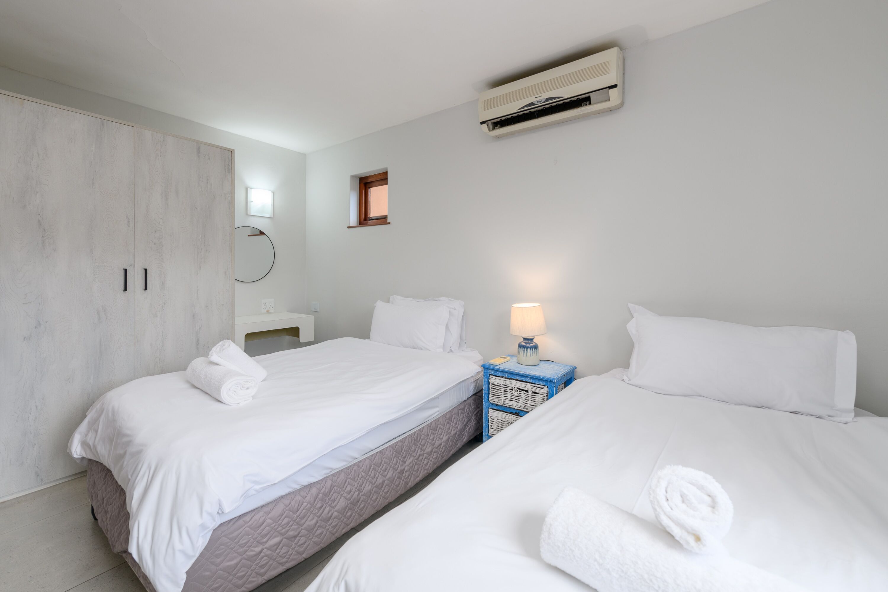 Quarto de hotel com duas camas, um guarda-roupa, uma mesinha com abajur e um aparelho de ar condicionado.