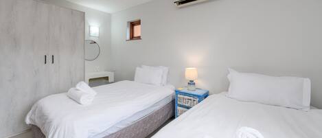 Quarto de hotel com duas camas, um guarda-roupa, uma mesinha com abajur e um aparelho de ar condicionado.