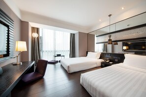 Quarto de hotel com duas camas, uma escrivaninha com luminária, uma cadeira, uma TV e uma janela grande com cortinas.