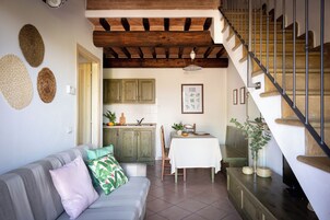 Uma sala de estar aconchegante com um sofá cinza, uma mesa de jantar com toalha branca e uma área de cozinha com armários de madeira e painel de azulejos.